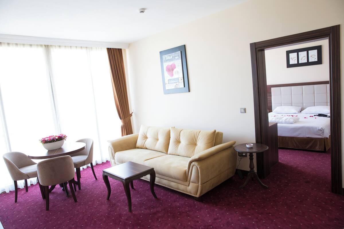 Quarto de hotel com um sofá bege, uma mesa redonda pequena, um espaço de refeições com cadeiras, uma cama com roupa de cama branca e uma moldura com uma imagem na parede.