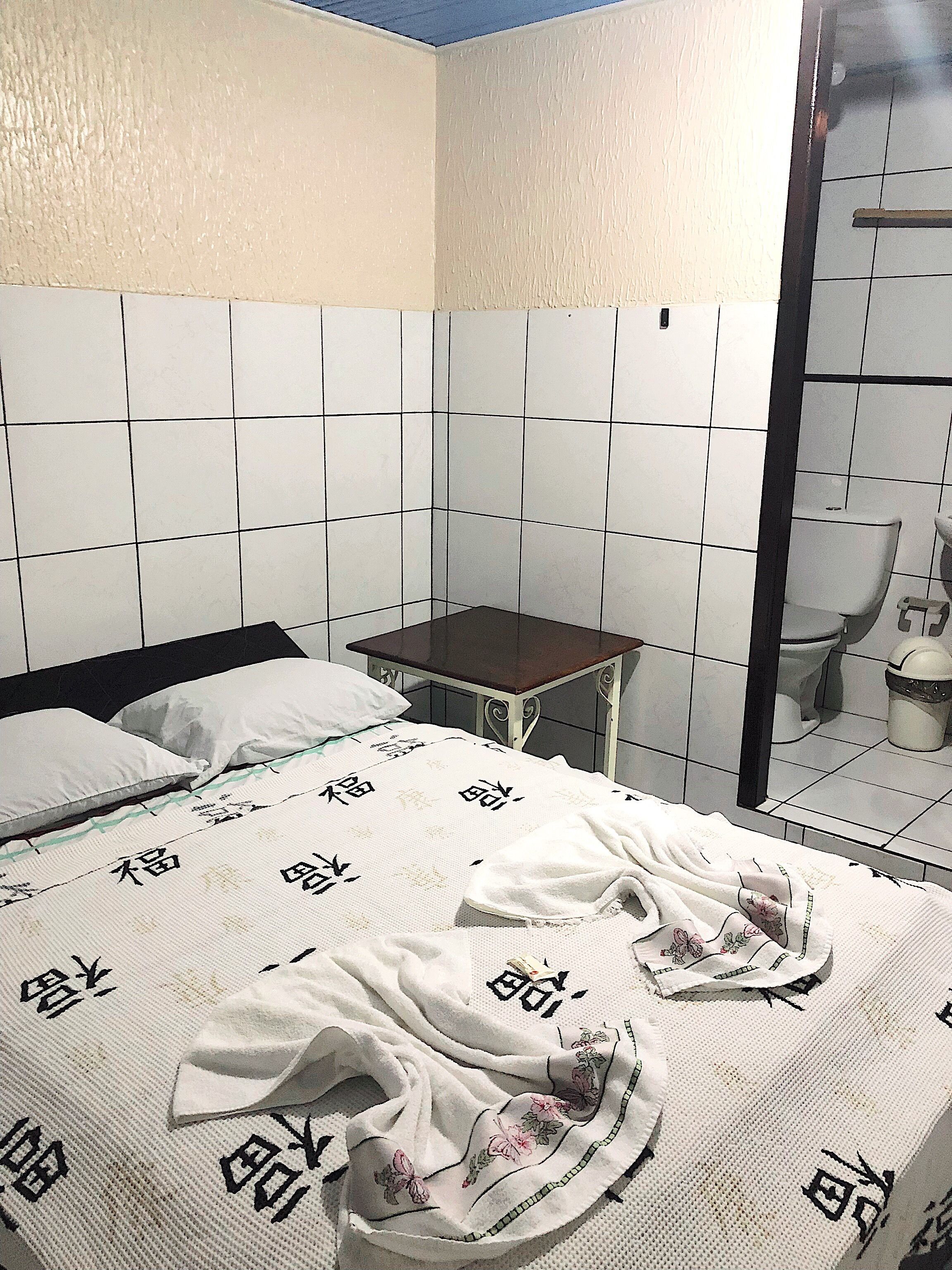 Um quarto pequeno com azulejos, uma cama, uma mesinha e um vaso sanitário.