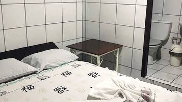 Um quarto pequeno com azulejos, uma cama, uma mesinha e um vaso sanitário.