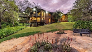 Um edifício de dois andares com varanda, rodeado por um jardim ajardinado com diversas plantas e um banco.