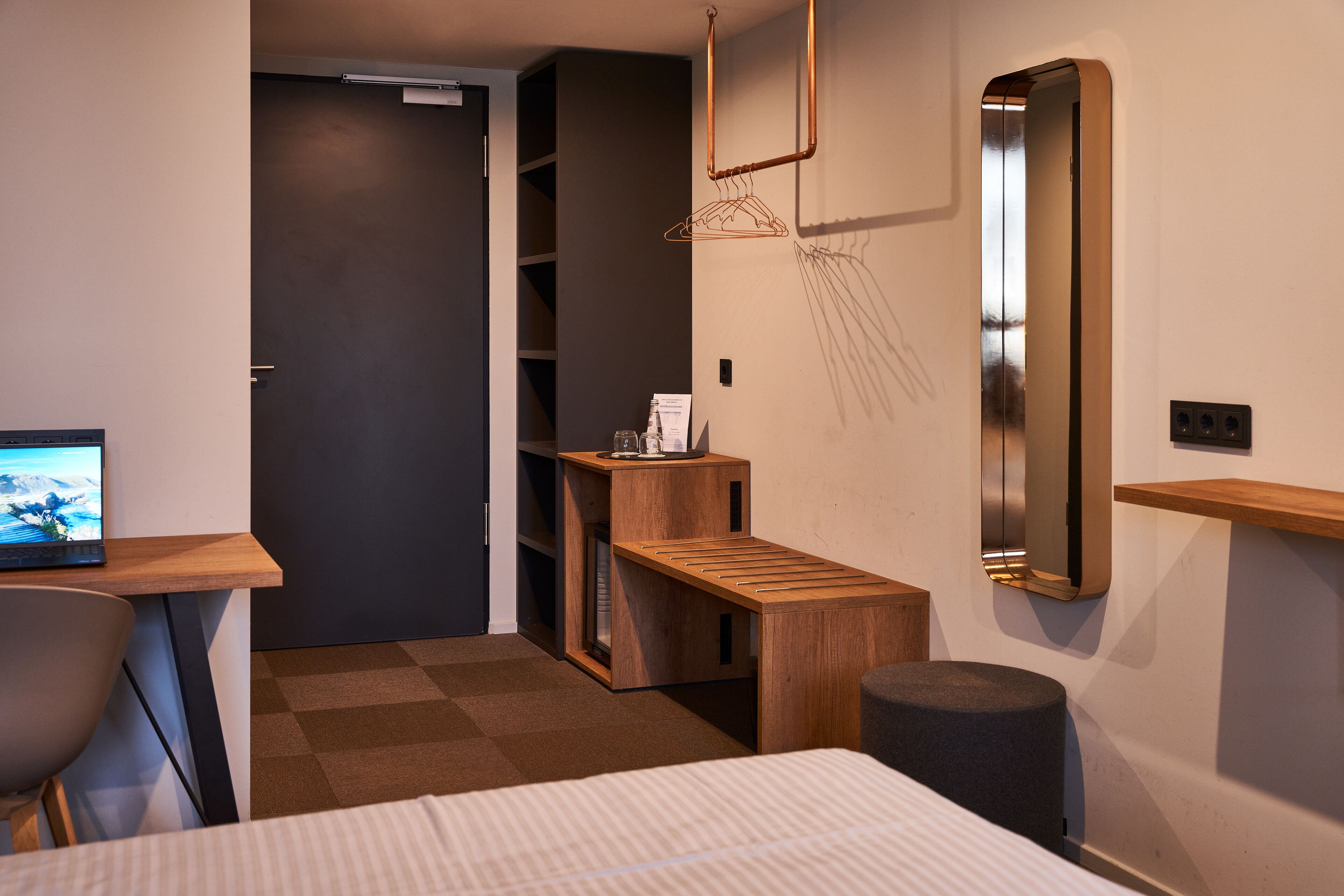 Quarto de hotel moderno com uma cama, uma escrivaninha com um laptop, um banco de madeira, um espelho e um guarda-roupa.