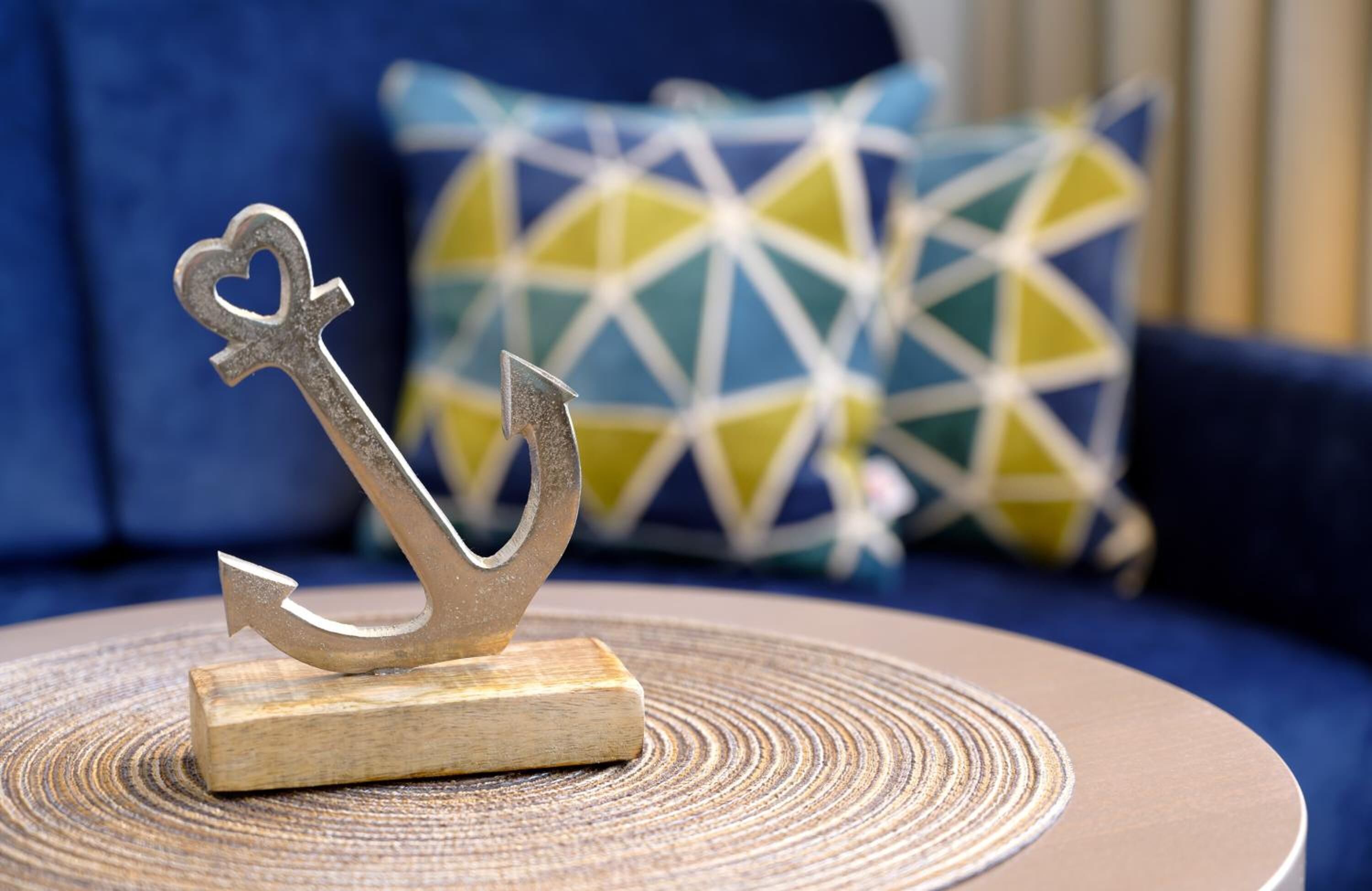 Uma âncora decorativa sobre um bloco de madeira, colocada sobre uma mesa redonda com textura, com um sofá azul e uma almofada estampada ao fundo.