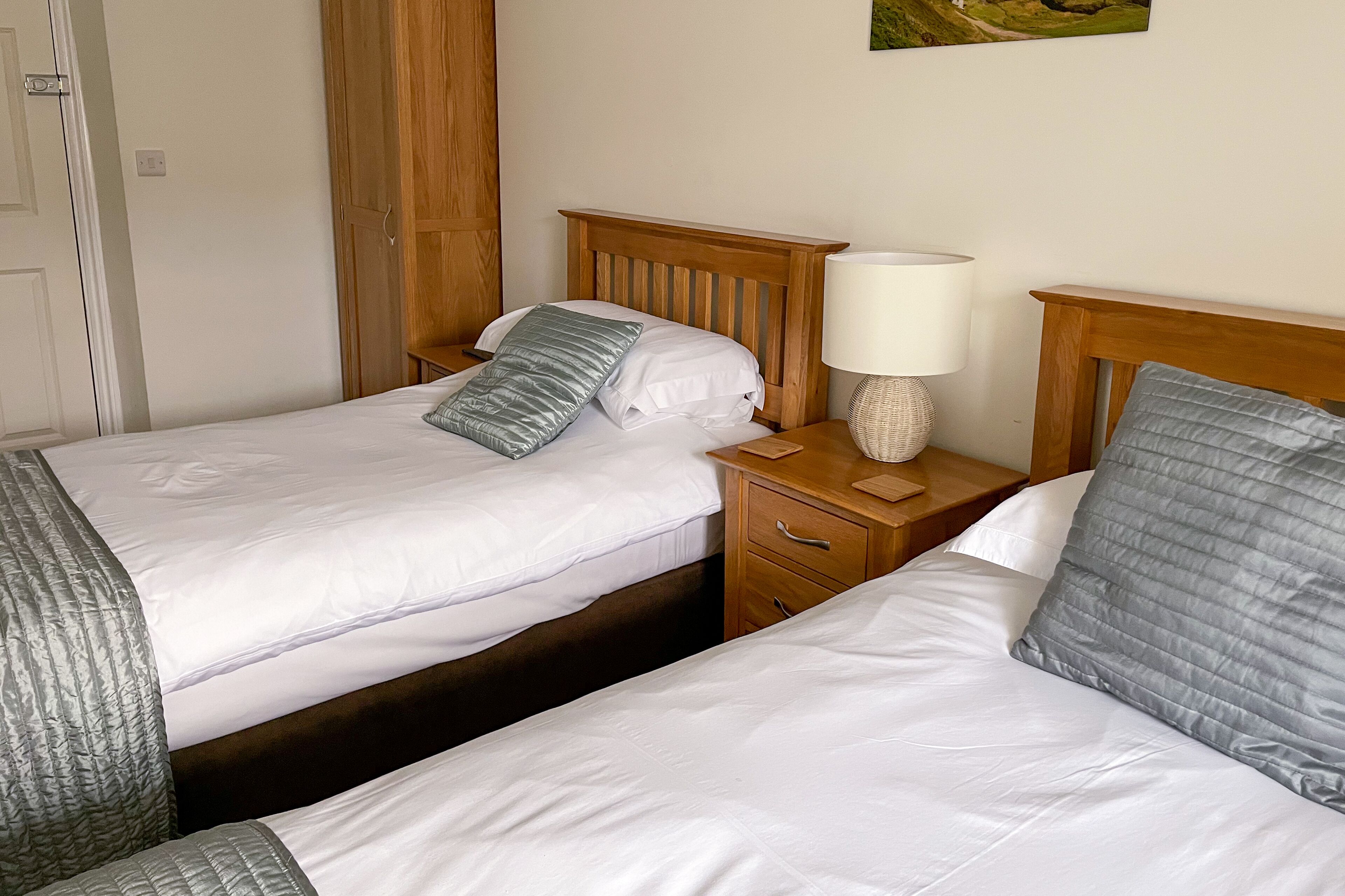 Uma cama de casal com lençóis brancos e cabeceira de madeira, uma mesa de cabeceira com abajur e um guarda-roupa de madeira.