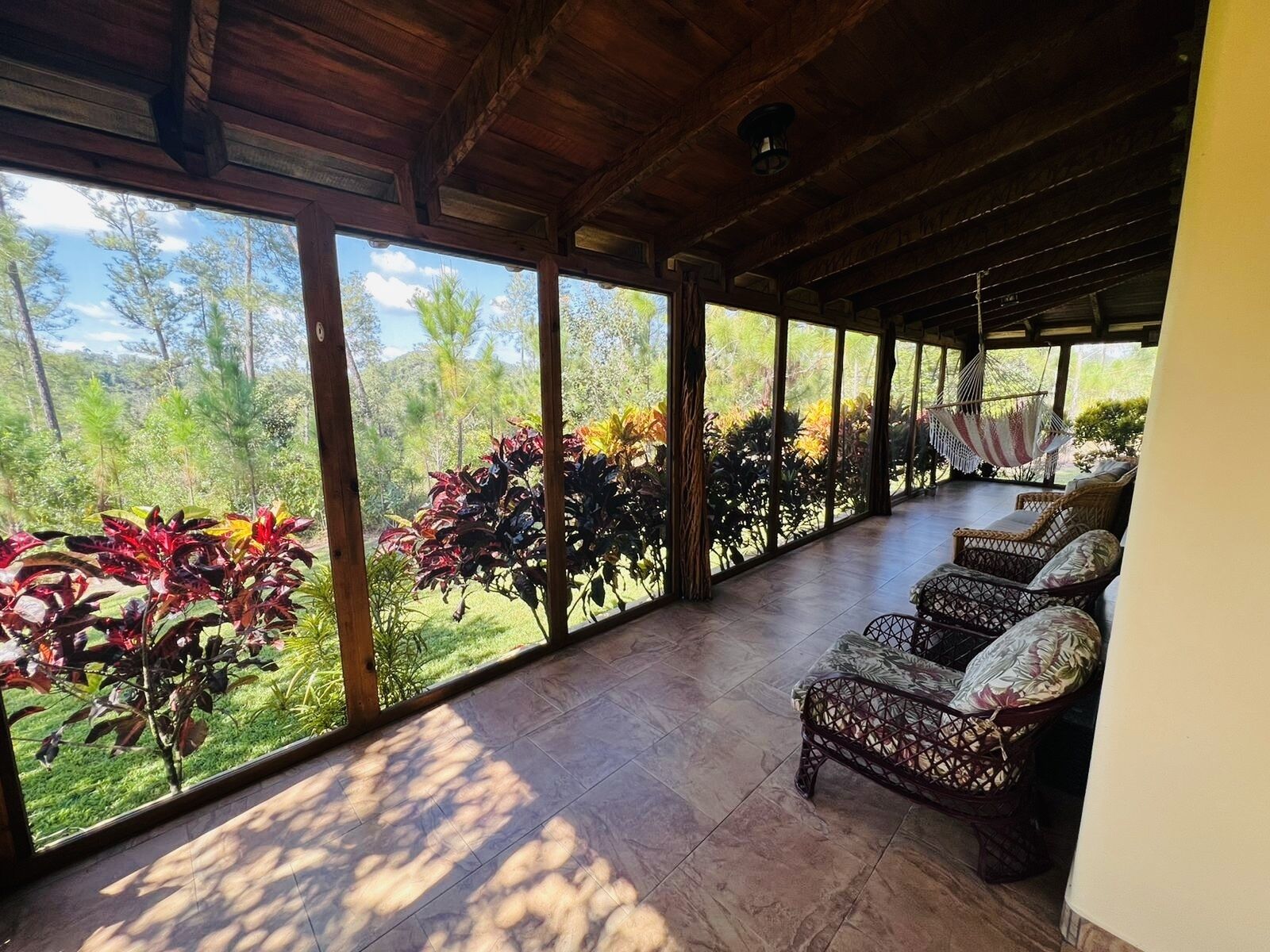 Uma área de estar coberta ao ar livre com móveis de vime e uma rede, com vista para um jardim com diversas plantas e árvores.