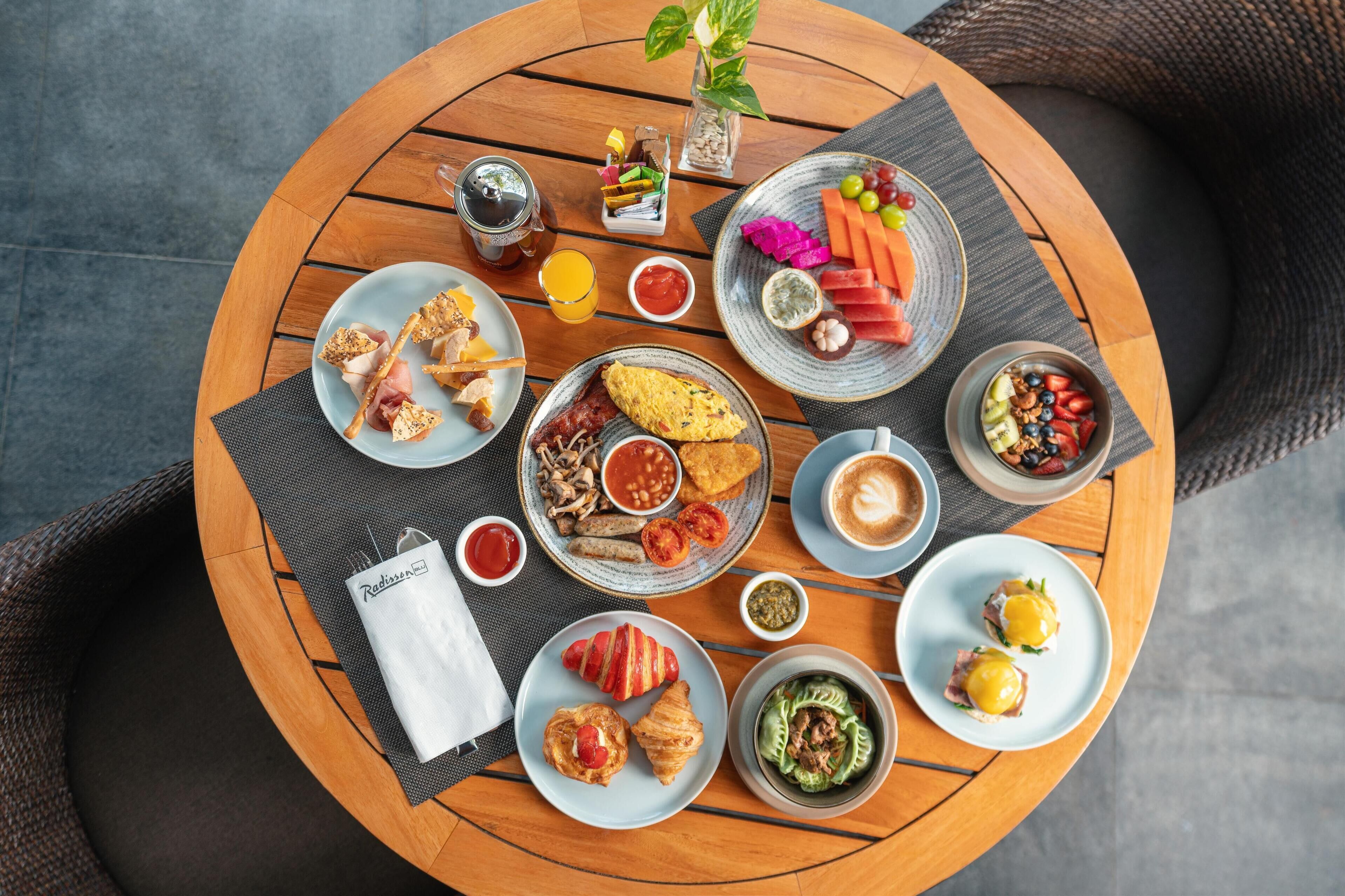 Mesa de madeira redonda com diversos itens de café da manhã, como croissants, ovos e frutas.