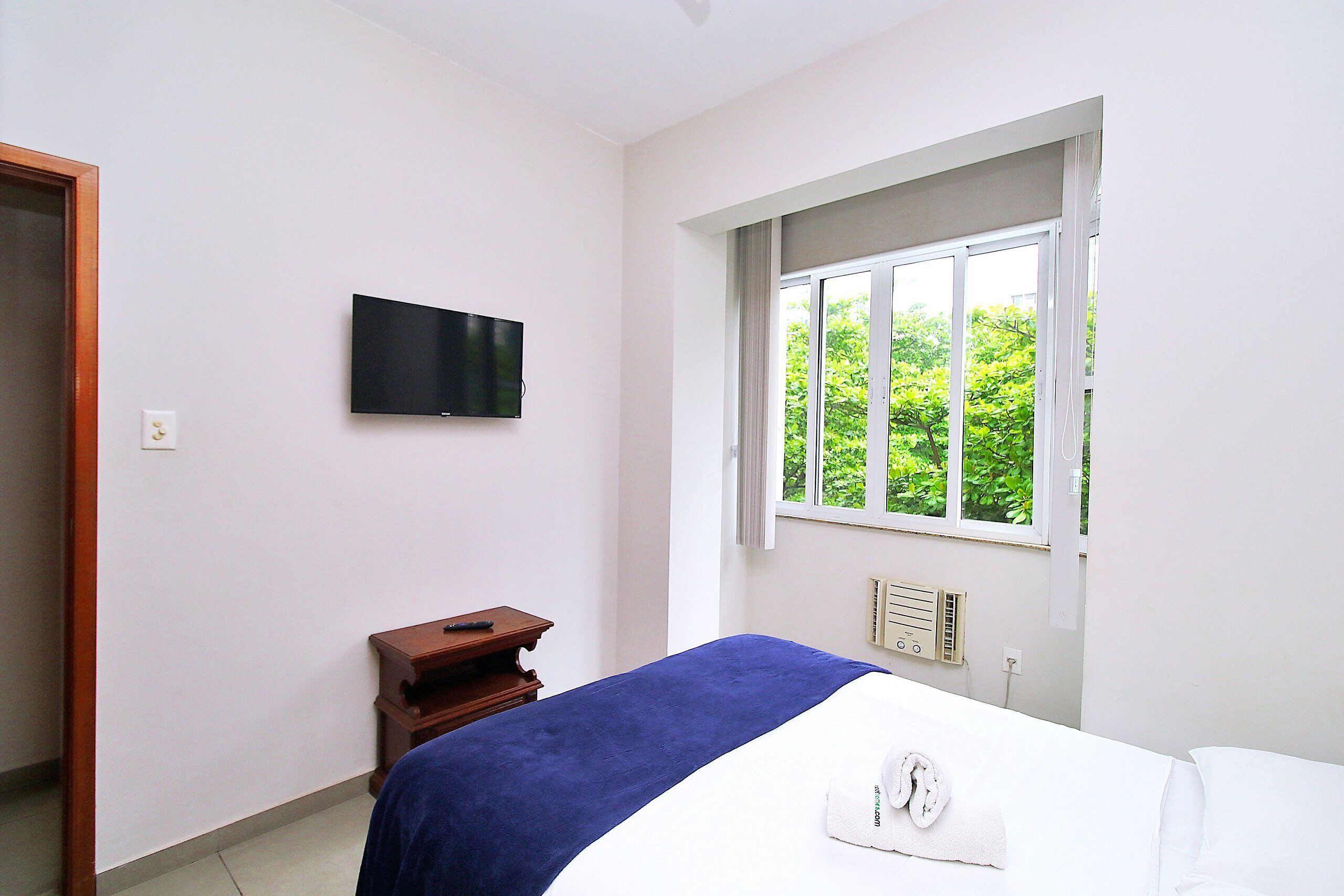Quarto com cama, televisão presa na parede, janela com vista para área verde e uma pequena mesa de cabeceira de madeira.