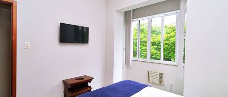 Quarto com cama, televisão presa na parede, janela com vista para área verde e uma pequena mesa de cabeceira de madeira.
