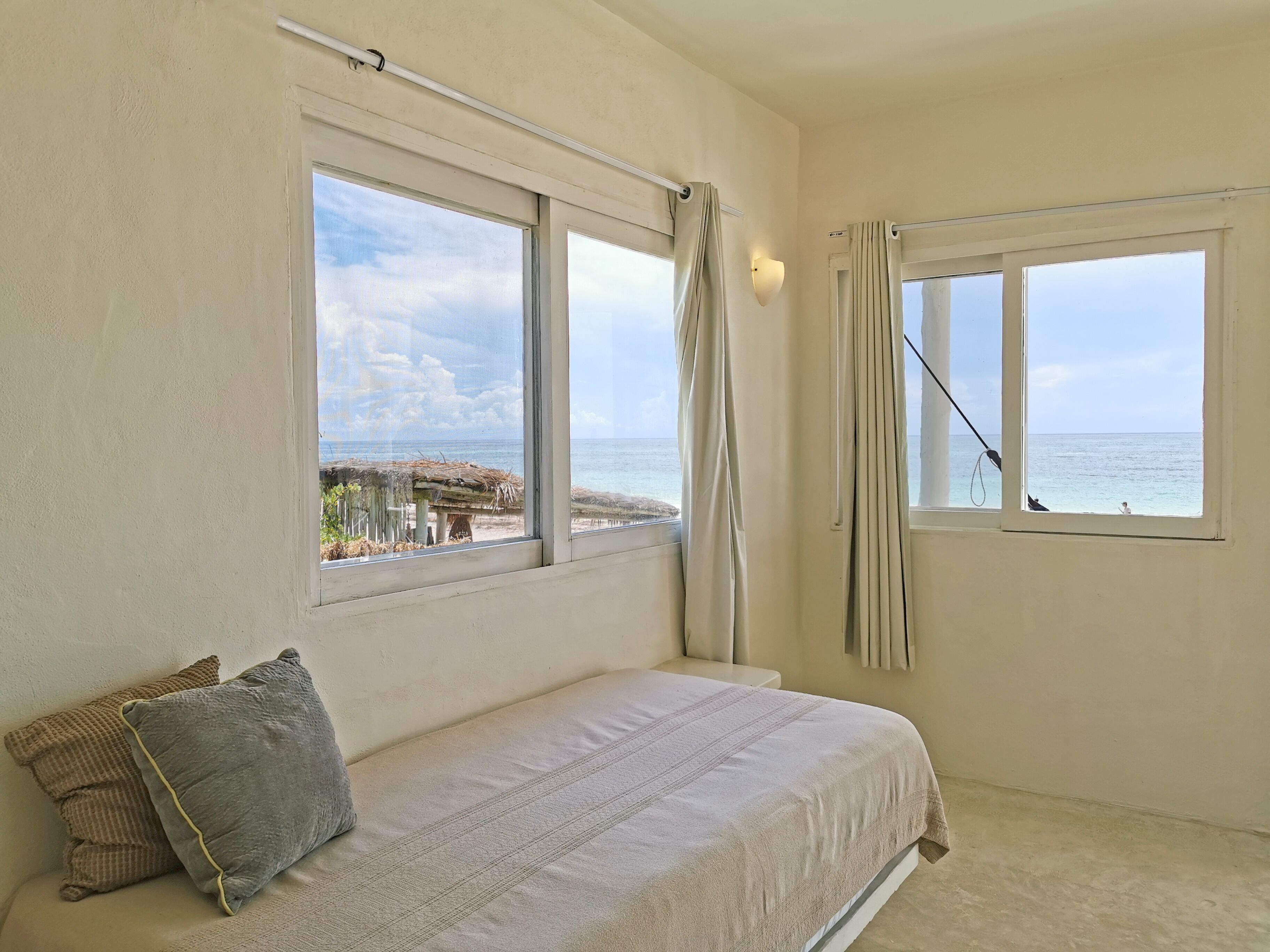 Quarto com cama, duas janelas com cortinas, vista para a praia e uma mesa pequena.
