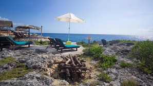 Uma praia com cadeiras de praia, uma estrutura com telhado de palha e um primeiro plano rochoso.