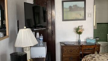 Quarto com cama, escrivaninha com luminária, televisão, cafeteira, garrafa de água, quadro e cômoda.