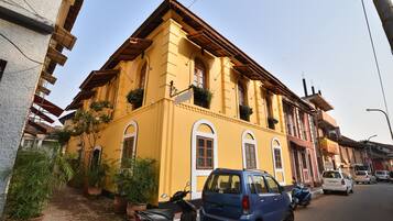 Um edifício amarelo com telhado em telhas, vasos de plantas e veículos estacionados.