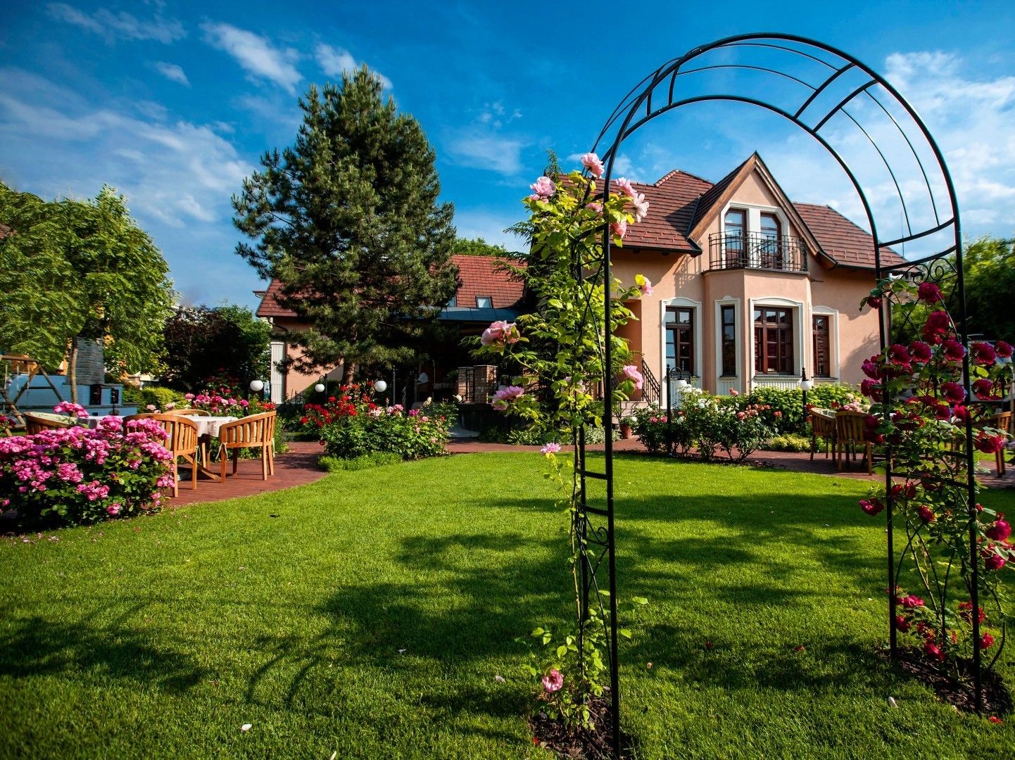 Uma casa com jardim bem cuidado, uma pérgula coberta de rosas trepadeiras e uma área de refeições com mobiliário ao ar livre.
