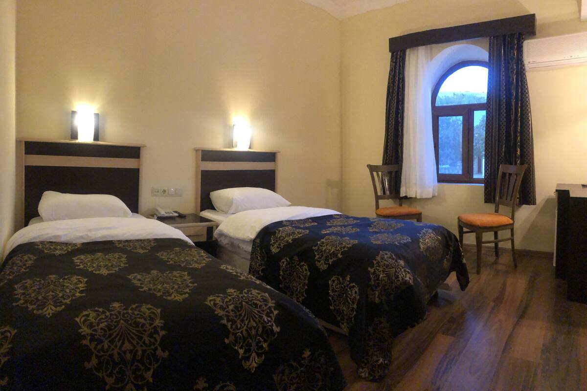 Quarto de hotel com duas camas, uma cadeira, uma escrivaninha e uma janela com cortinas.