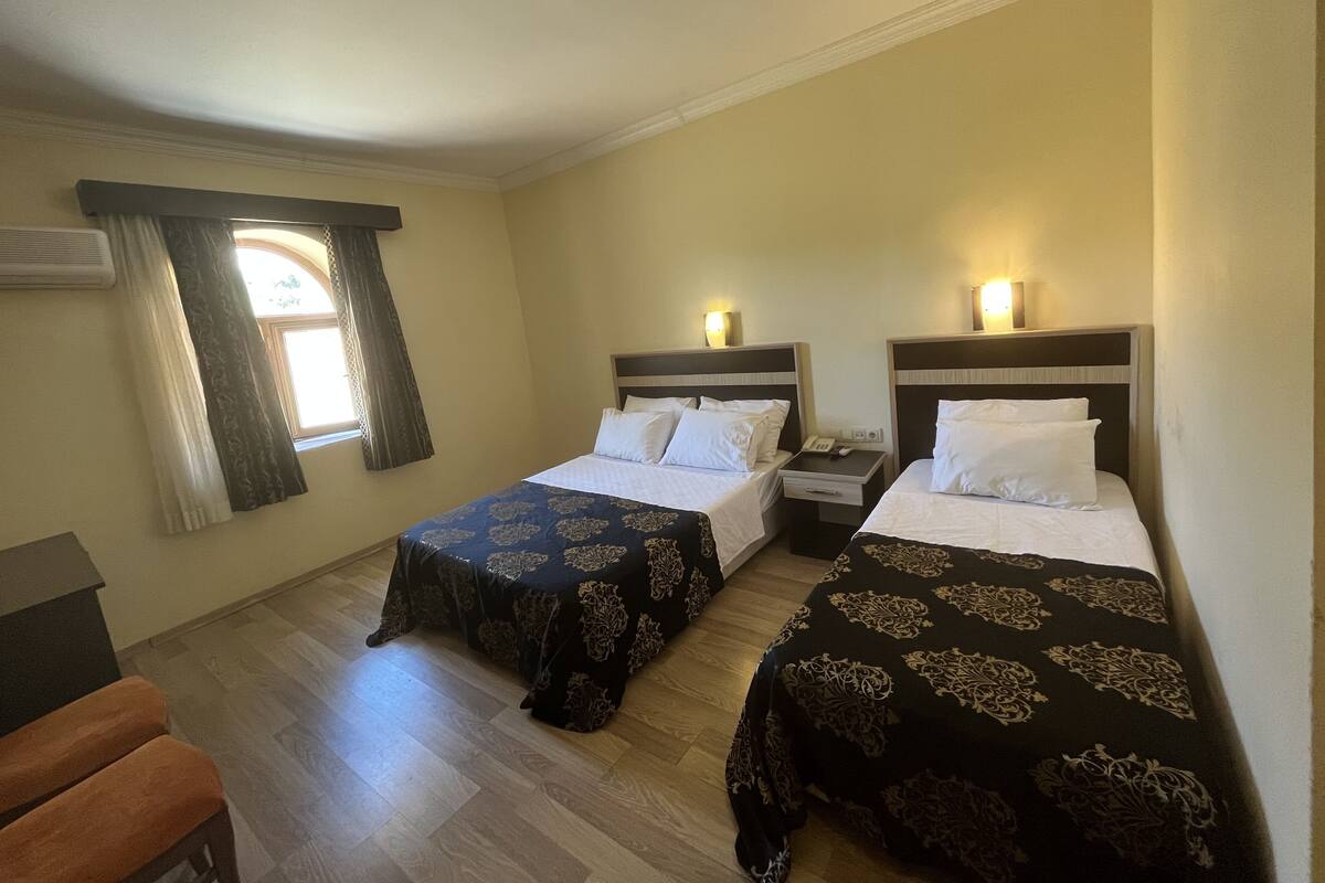 Quarto de hotel com duas camas, cada uma com cabeceira e mesas de cabeceira. Há uma janela com cortinas, uma cadeira e um aparelho de ar condicionado na parede.