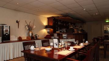 Uma sala de jantar com uma longa mesa de madeira posta para seis pessoas, rodeada por cadeiras. Há também um espaço bar com prateleiras de garrafas e copos. Uma máquina de café e um vaso com flores também estão visíveis.