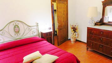 Quarto com colcha vermelha, cómoda de madeira, espelho e um vaso com flores.