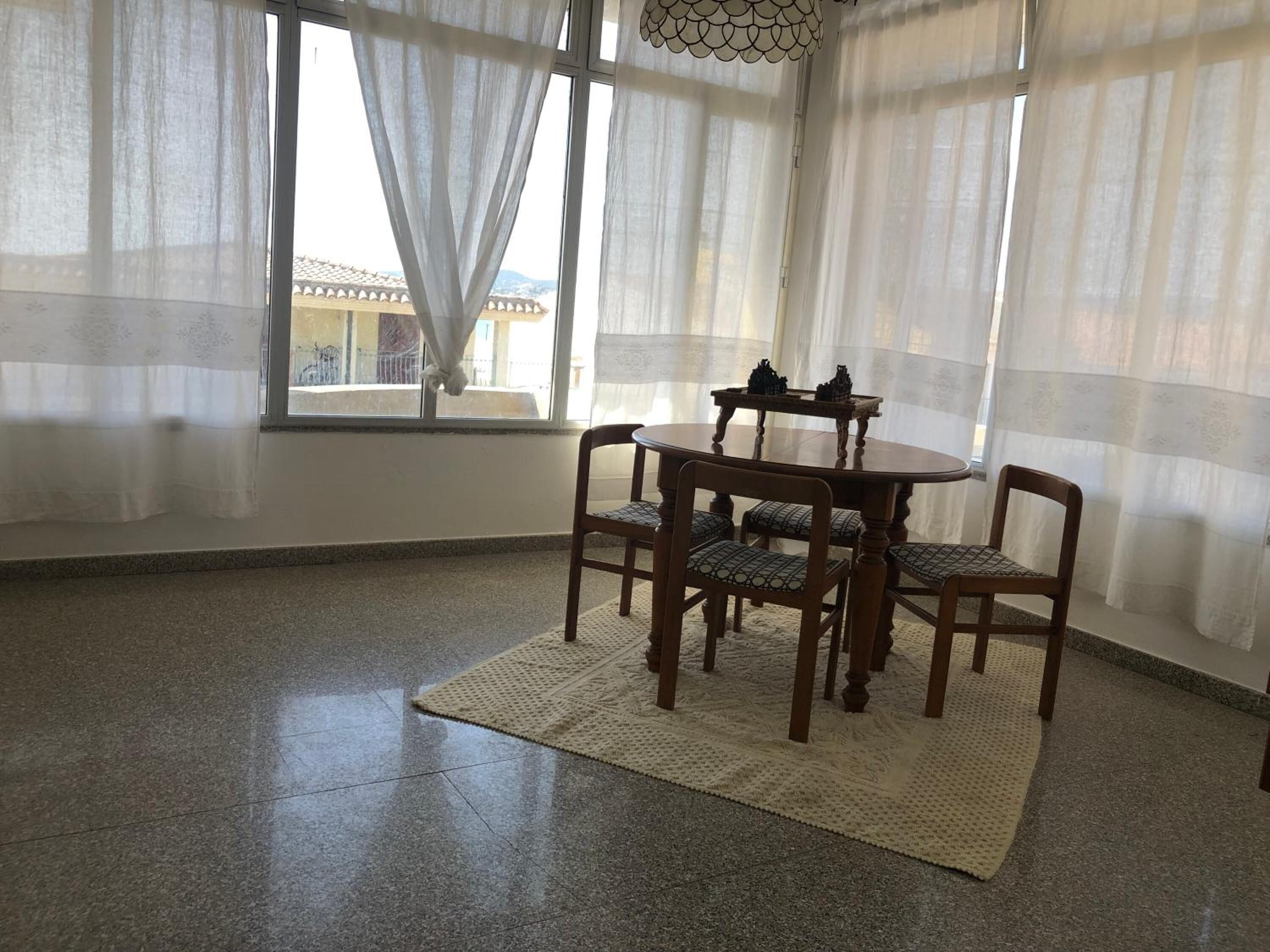 Um quarto com uma mesa de madeira redonda e quatro cadeiras, um tapete tecido e cortinas leves.