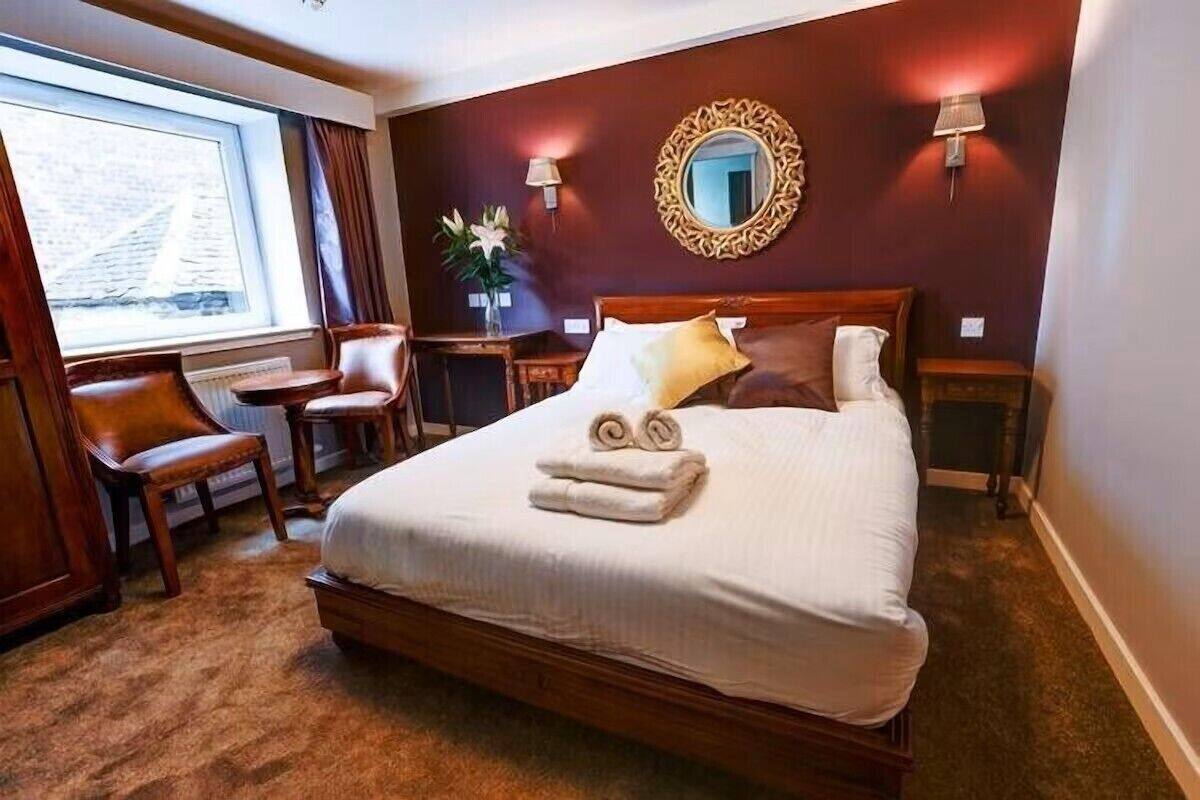 Quarto de hotel com uma cama grande, duas mesas de cabeceira, uma cadeira, uma janela com cortinas, um espelho e um vaso de planta.