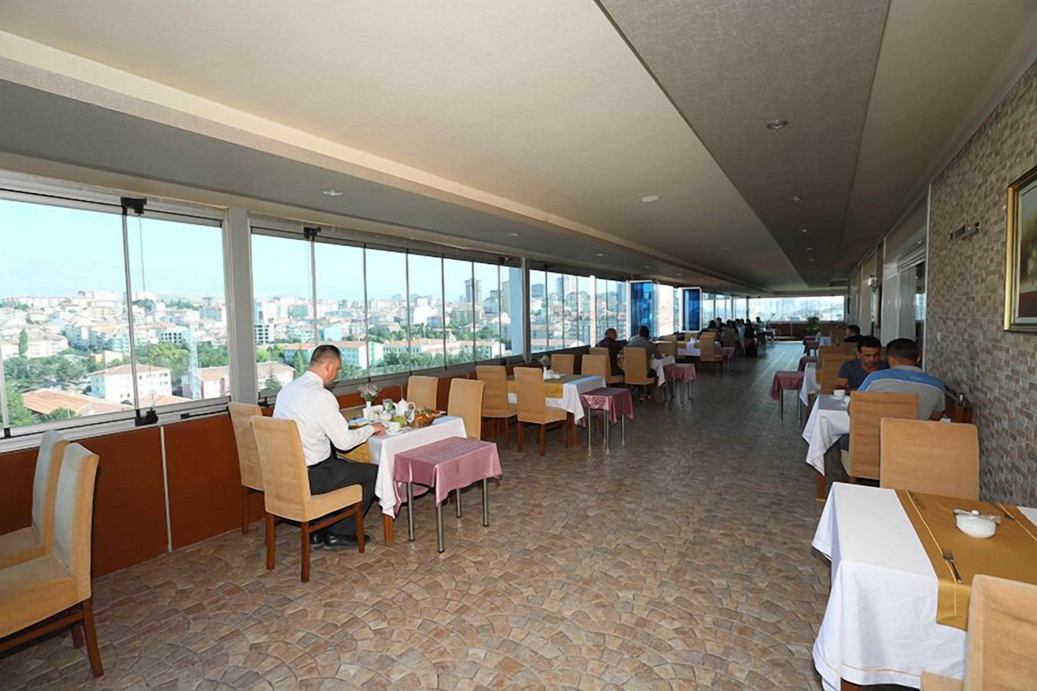 Um restaurante espaçoso com pé-direito alto e grandes janelas que oferecem vista da cidade. As mesas estão bem organizadas com cadeiras, e o chão é revestido de azulejos.