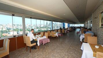 Um restaurante espaçoso com pé-direito alto e grandes janelas que oferecem vista da cidade. As mesas estão bem organizadas com cadeiras, e o chão é revestido de azulejos.