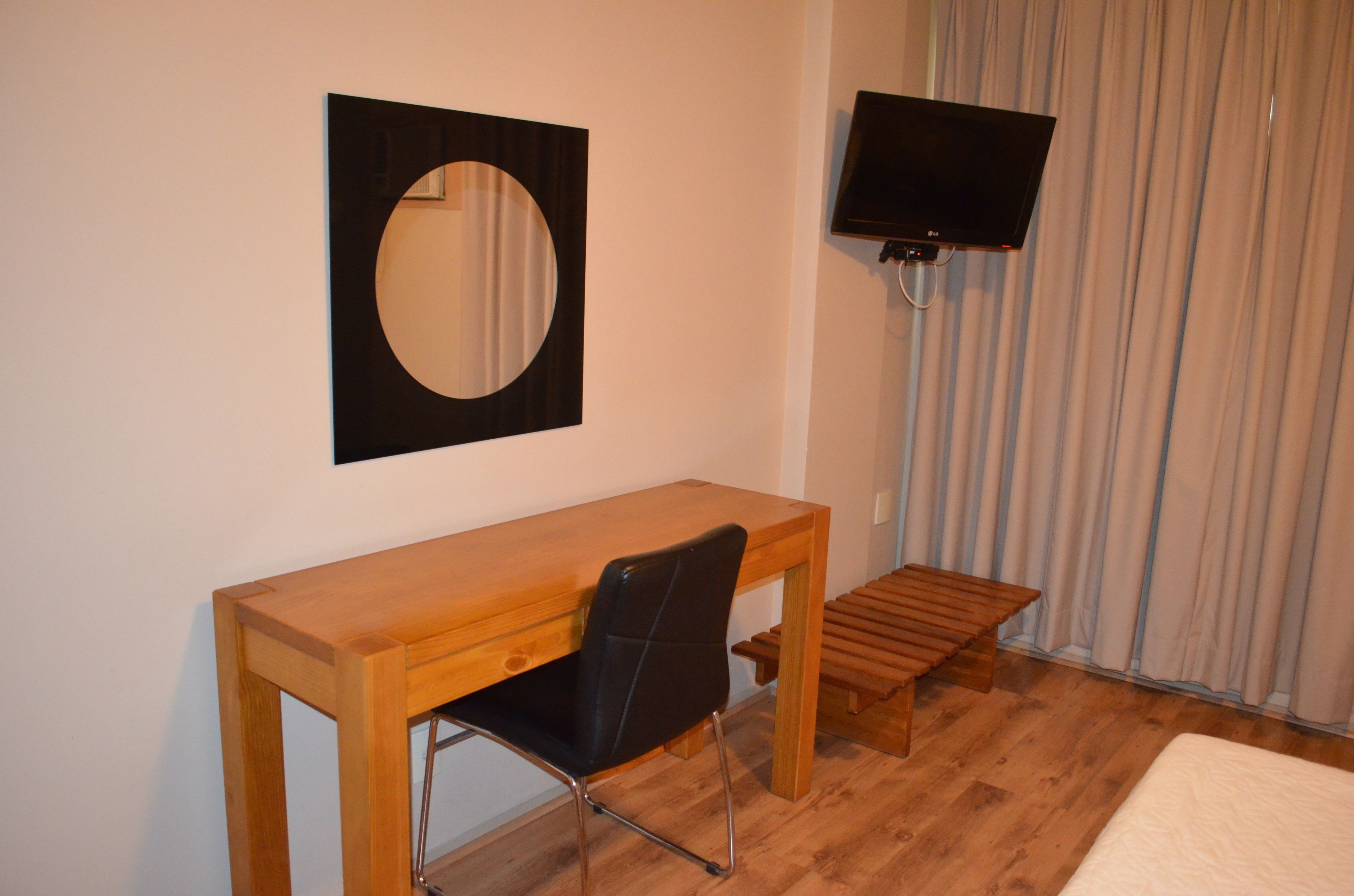 Quarto com escrivaninha de madeira, cadeira preta, televisão fixada na parede e janela com cortinas.
