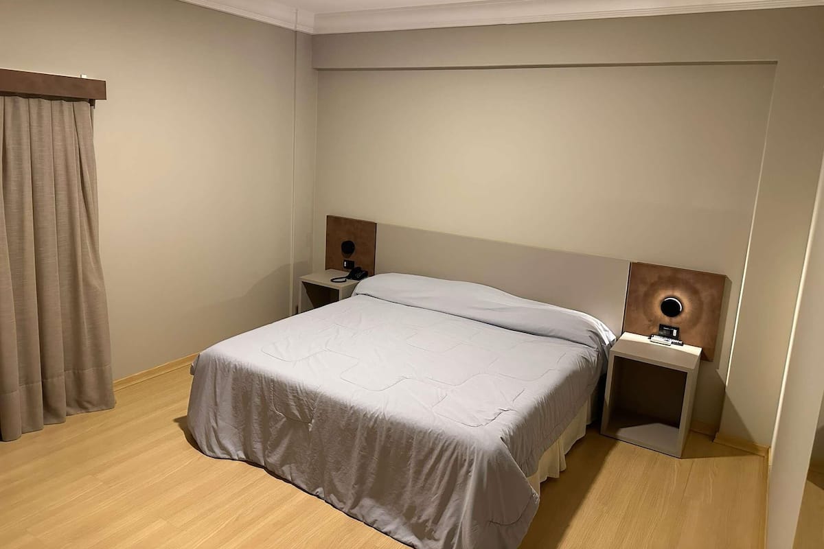 Quarto de hotel com cama, mesa de cabeceira com telefone, luminária na parede e cortina.