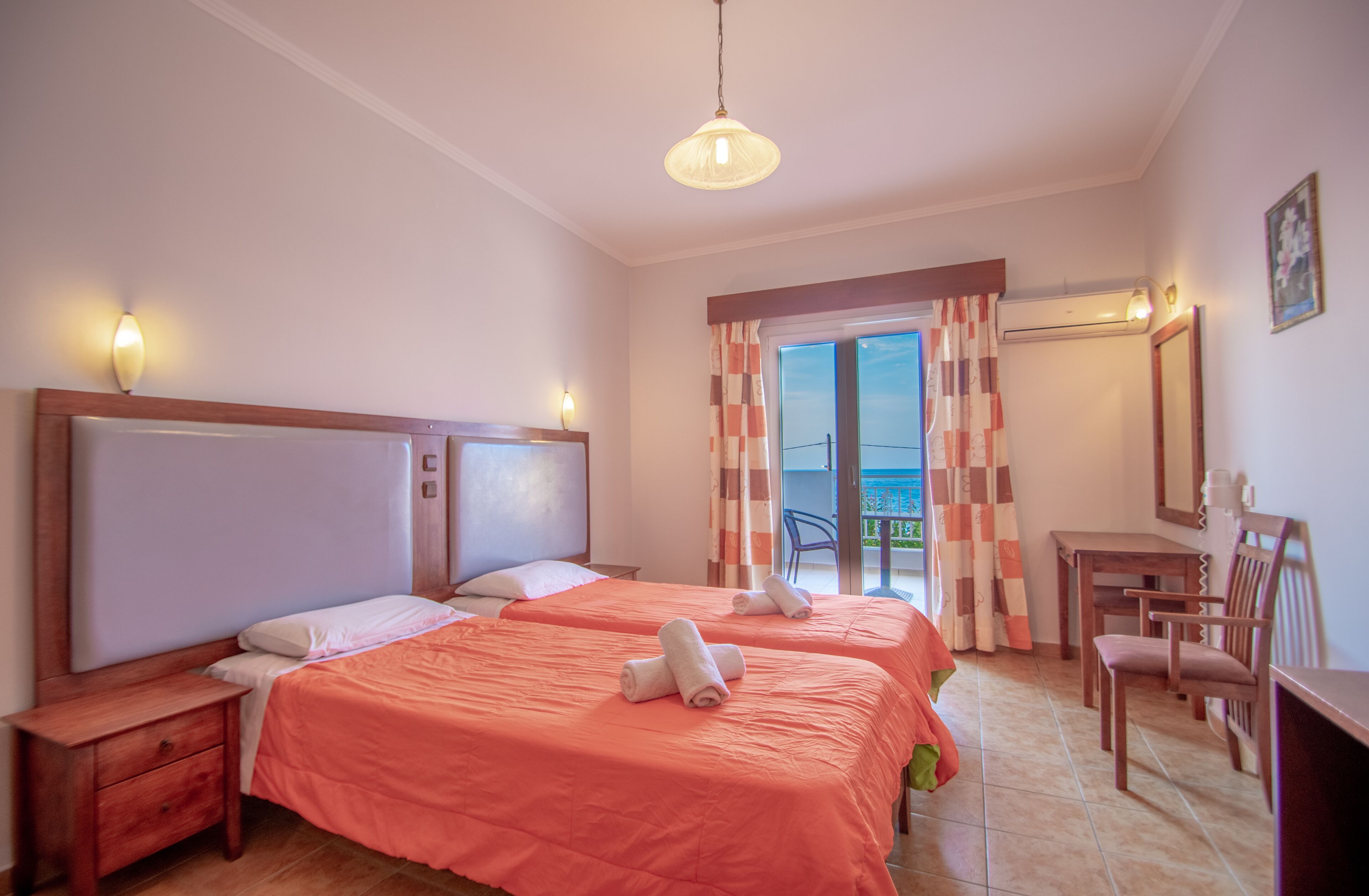 Quarto de hotel com duas camas, uma escrivaninha, uma cadeira e uma varanda com vista.