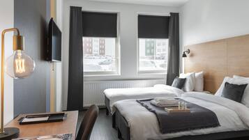 Quarto de hotel com duas camas, uma secretária com abajur, televisão e vista da rua por grandes janelas.