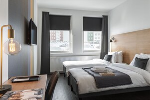 Quarto de hotel com duas camas, uma secretária com abajur, televisão e vista da rua por grandes janelas.