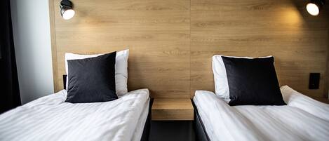 Duas camas de solteiro com roupa de cama branca e almofadas pretas, uma cabeceira de madeira e duas luzes fixadas na parede.