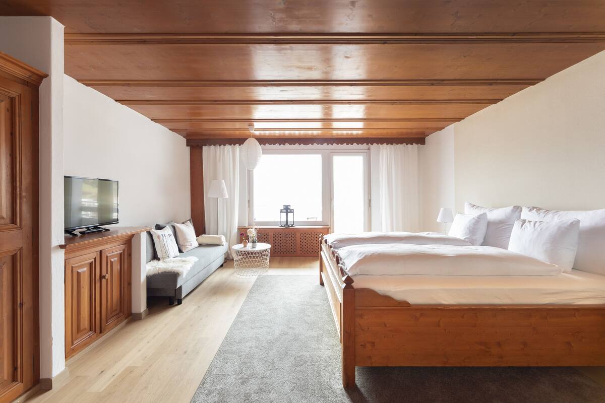 Quarto com teto de madeira, uma cama grande, televisão, sofá e janela com cortinas.
