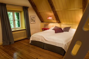 Quarto com piso de madeira, cama com roupa de cama branca, abajur vermelho e janela com cortinas.