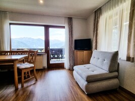 Um quarto com uma mesa de jantar de madeira, um sofá cinza, uma televisão em um armário de madeira e vista para montanhas por uma grande janela.