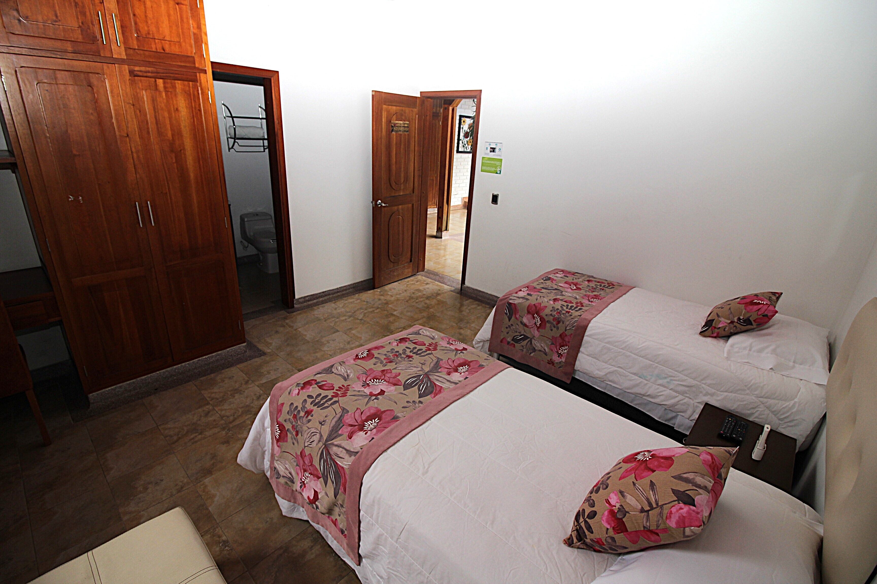 Quarto de hotel com duas camas, um guarda-roupa de madeira e um banheiro visível por uma porta aberta.