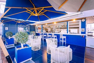 Uma área de bar com móveis de vime branco, paredes azuis e teto com vigas de madeira.