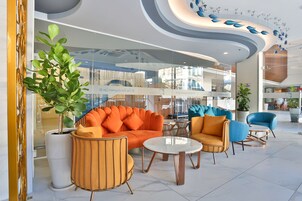 Um hall moderno com área de estar, com cadeiras laranja e azuis, uma mesa de centro branca e uma grande planta em vaso.