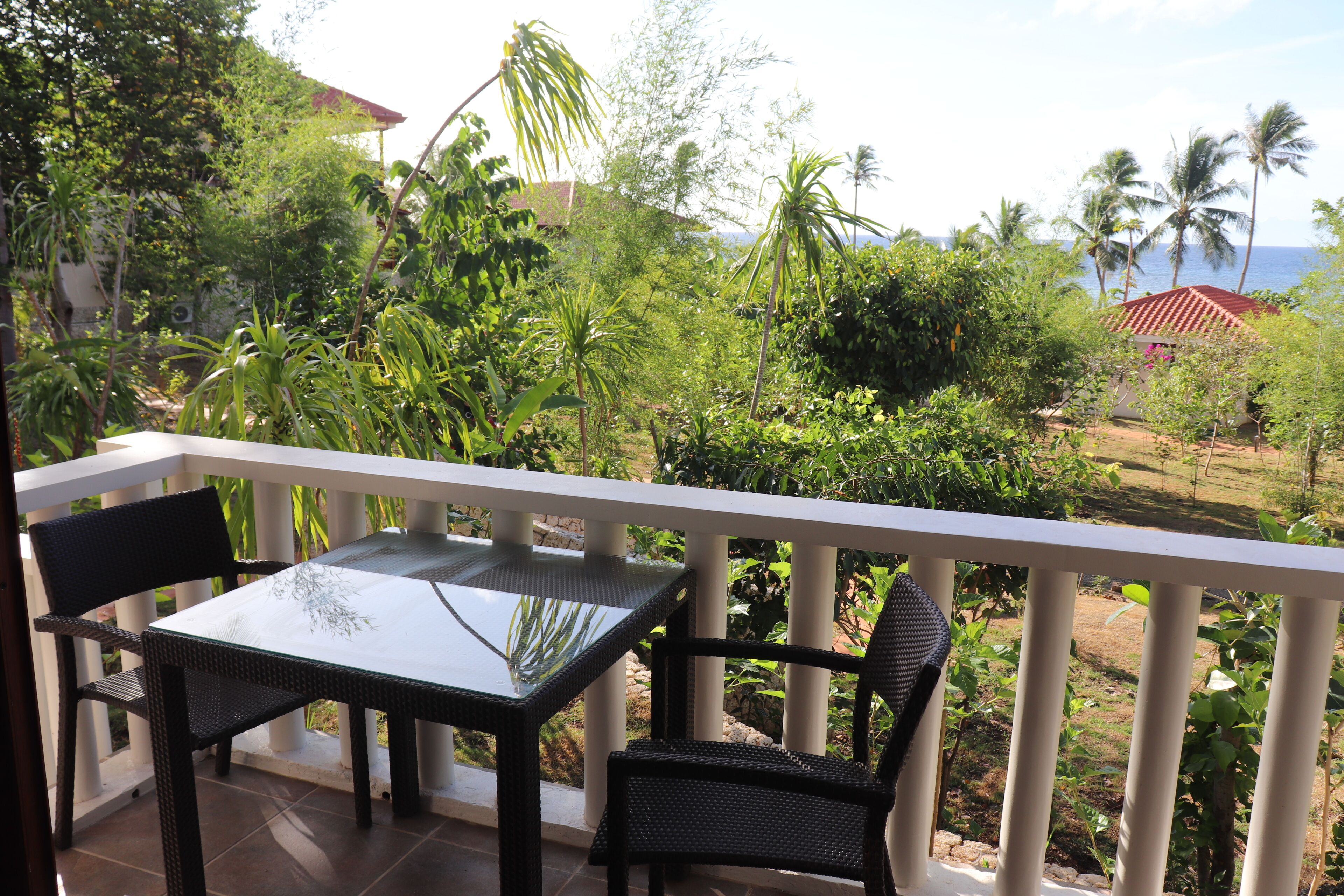 Uma varanda com mesa de vidro e duas cadeiras de vime, com vista para um jardim exuberante e o oceano.