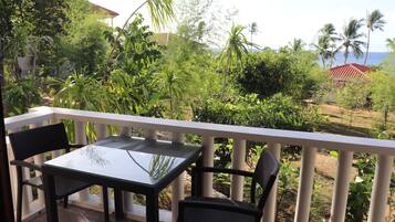 Uma varanda com mesa de vidro e duas cadeiras de vime, com vista para um jardim exuberante e o oceano.