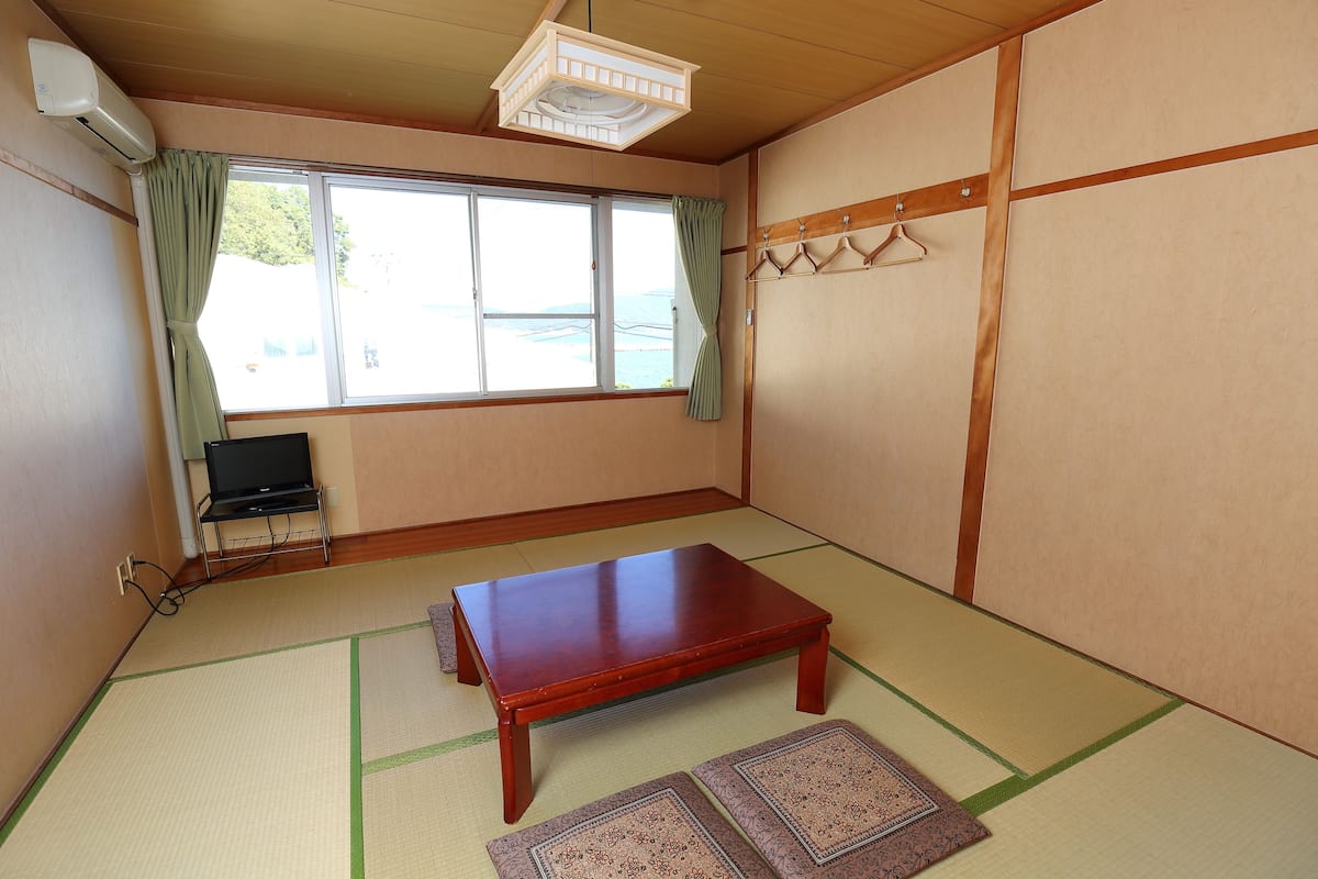 Um quarto japonês tradicional com tatames, uma mesa de madeira baixa, uma televisão em um suporte e vista para o exterior por uma grande janela.