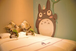 Quarto de hotel com uma cama, apresentando um bichinho de pelúcia do Totoro e um mural do Totoro na parede.