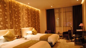 Quarto de hotel com duas camas, uma escrivaninha, uma cadeira e uma janela grande com cortinas.