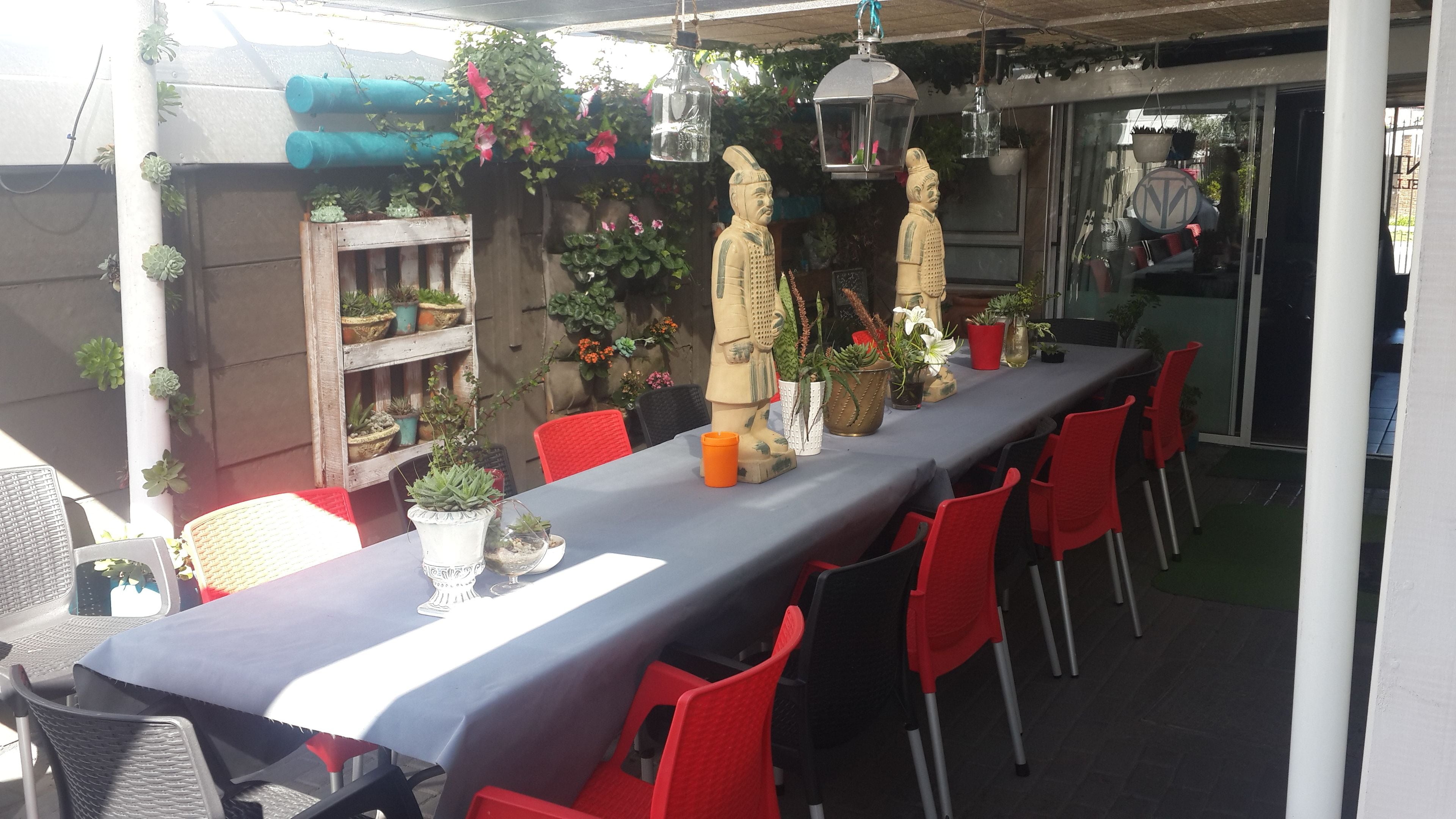 Uma área de refeições ao ar livre com uma mesa comprida, cadeiras vermelhas e diversos vasos de plantas.