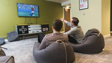Duas pessoas sentadas em sacos de feijão numa sala com uma TV mostrando um jogo esportivo, um armário com gavetas e uma porta ao fundo.