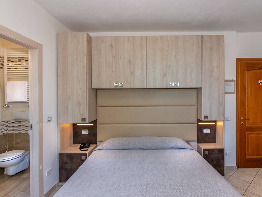 Quarto de hotel com cama, mesas de cabeceira, cabeceira de madeira, banheiro com vaso sanitário e pia, e uma porta com placa.