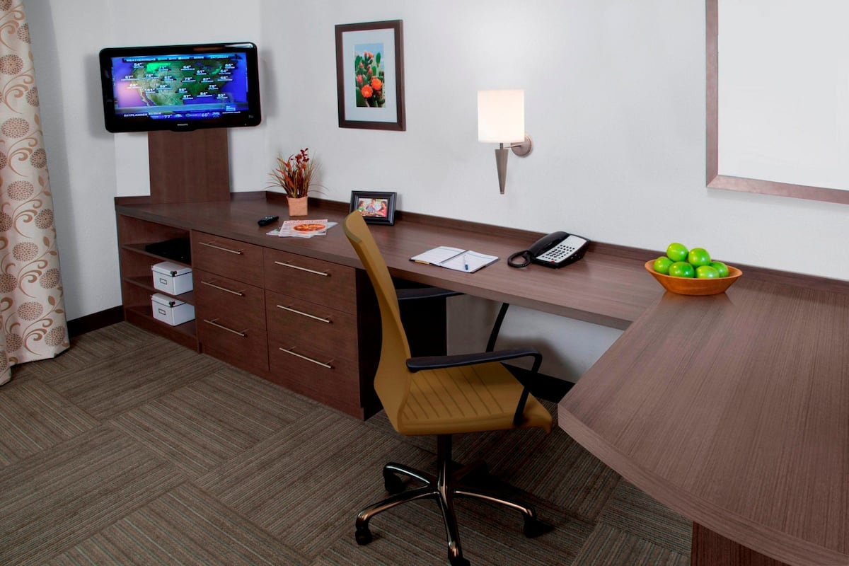 Escritório de hotel com uma mesa, cadeira, televisão, quadro emoldurado, luminária, telefone e uma tigela de frutas.