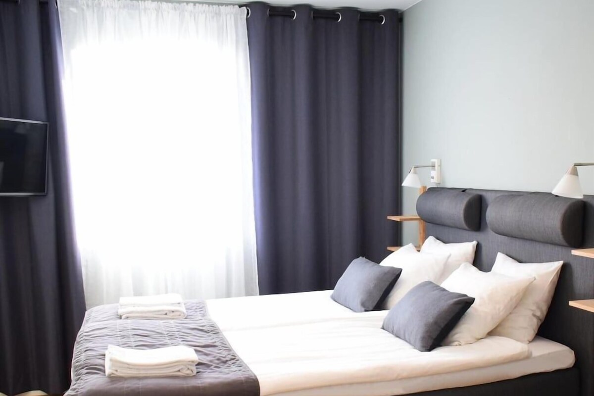 Quarto de hotel moderno com uma cama grande, uma TV de tela plana e uma mesa de cabeceira com luminárias.