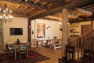Interior rústico com vigas de madeira aparentes, um candelabro, área de refeições com mobiliário de madeira e cozinha visível ao fundo.