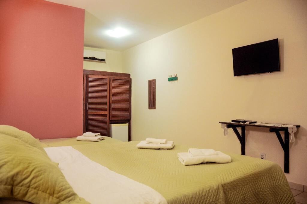 Quarto de hotel com uma cama, televisão na parede, uma mesinha e uma janela com persianas.