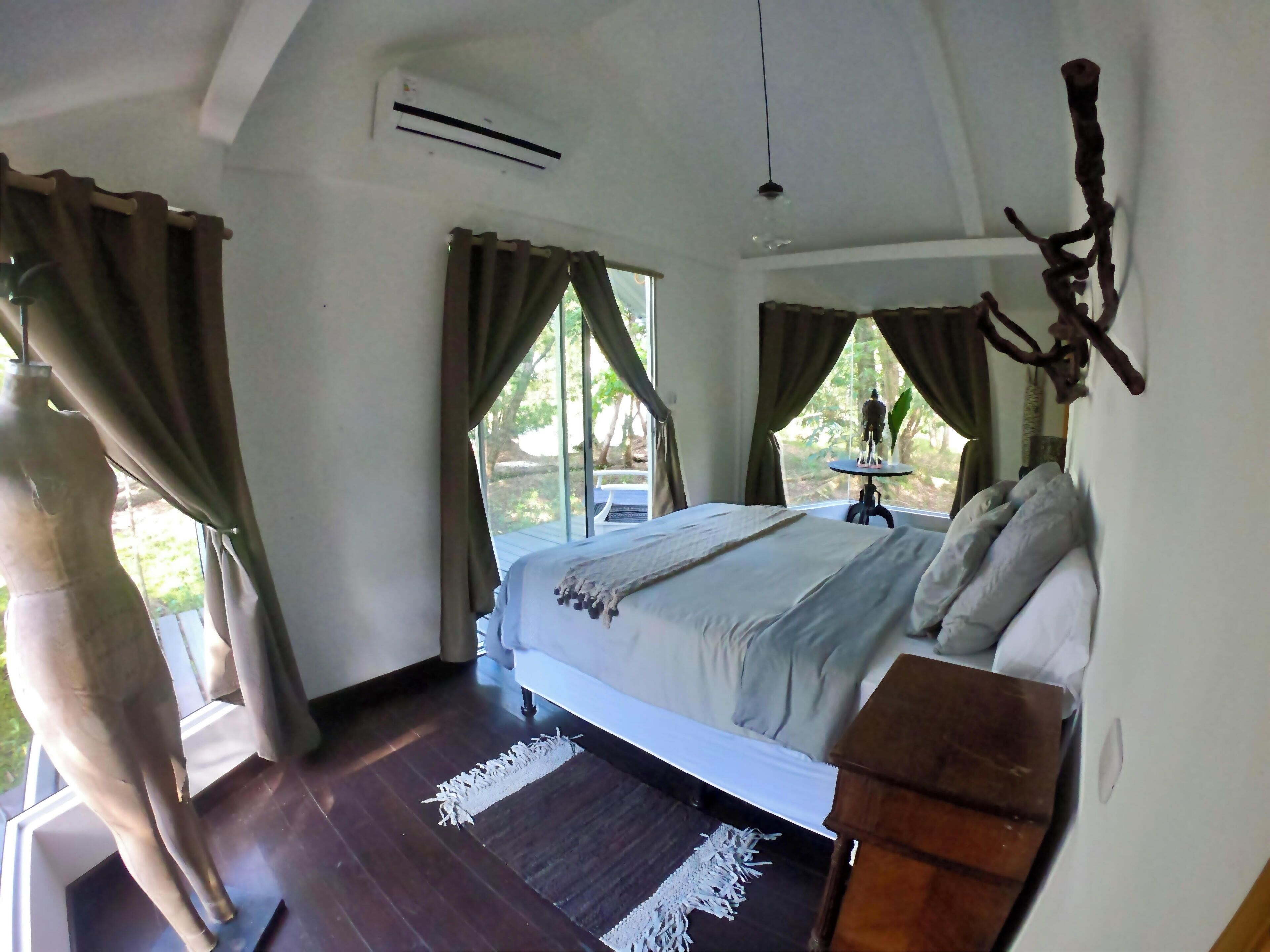 Quarto com cama, mesa de cabeceira de madeira, pequena mesa e luminária pendente.