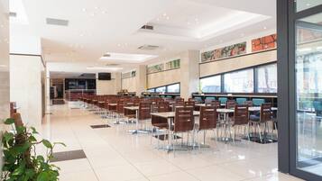 Uma cafeteria espaçosa com fileiras de mesas e cadeiras, uma janela grande com vista para o exterior, e um teto com iluminação embutida.